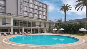 Piscina com espreguiçadeiras e guarda-sóis, rodeada por um edifício com varandas e palmeiras.