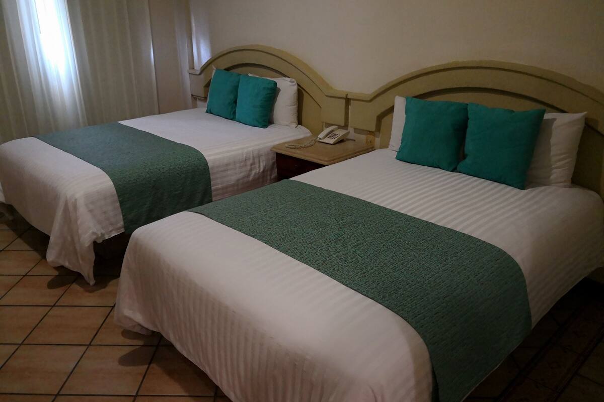 Duas camas de solteiro com roupa de cama verde e branca, uma mesinha de cabeceira com telefone e uma janela com cortinas.