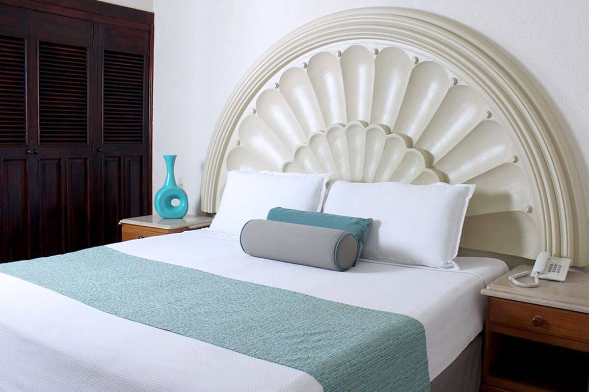 Uma cama com cabeceira branca, uma mesinha de cabeceira com um telefone e um criado-mudo com um vaso.