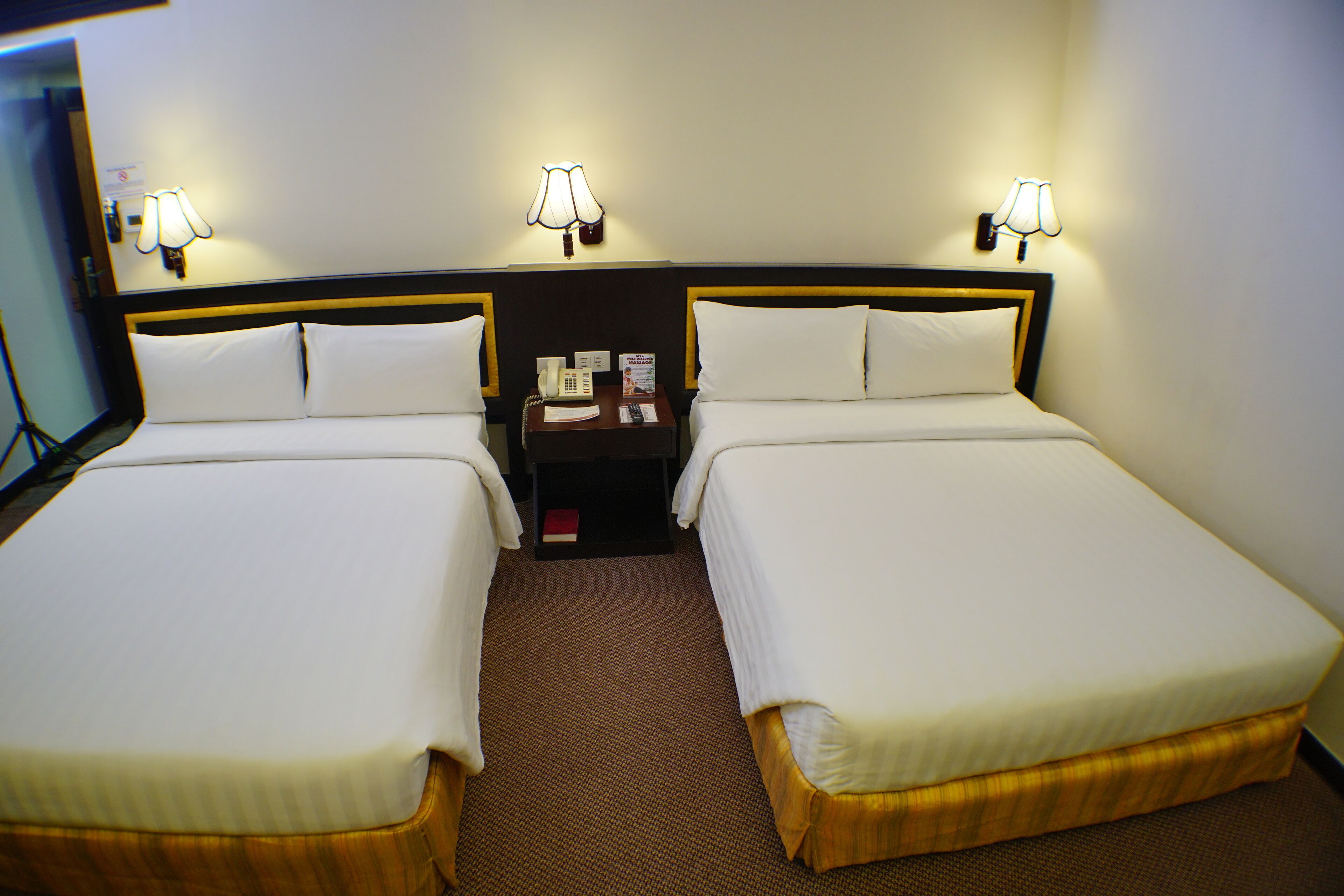 Quarto de hotel com duas camas de solteiro, uma mesinha de cabeceira com telefone e um livro, e luminárias fixadas na parede.