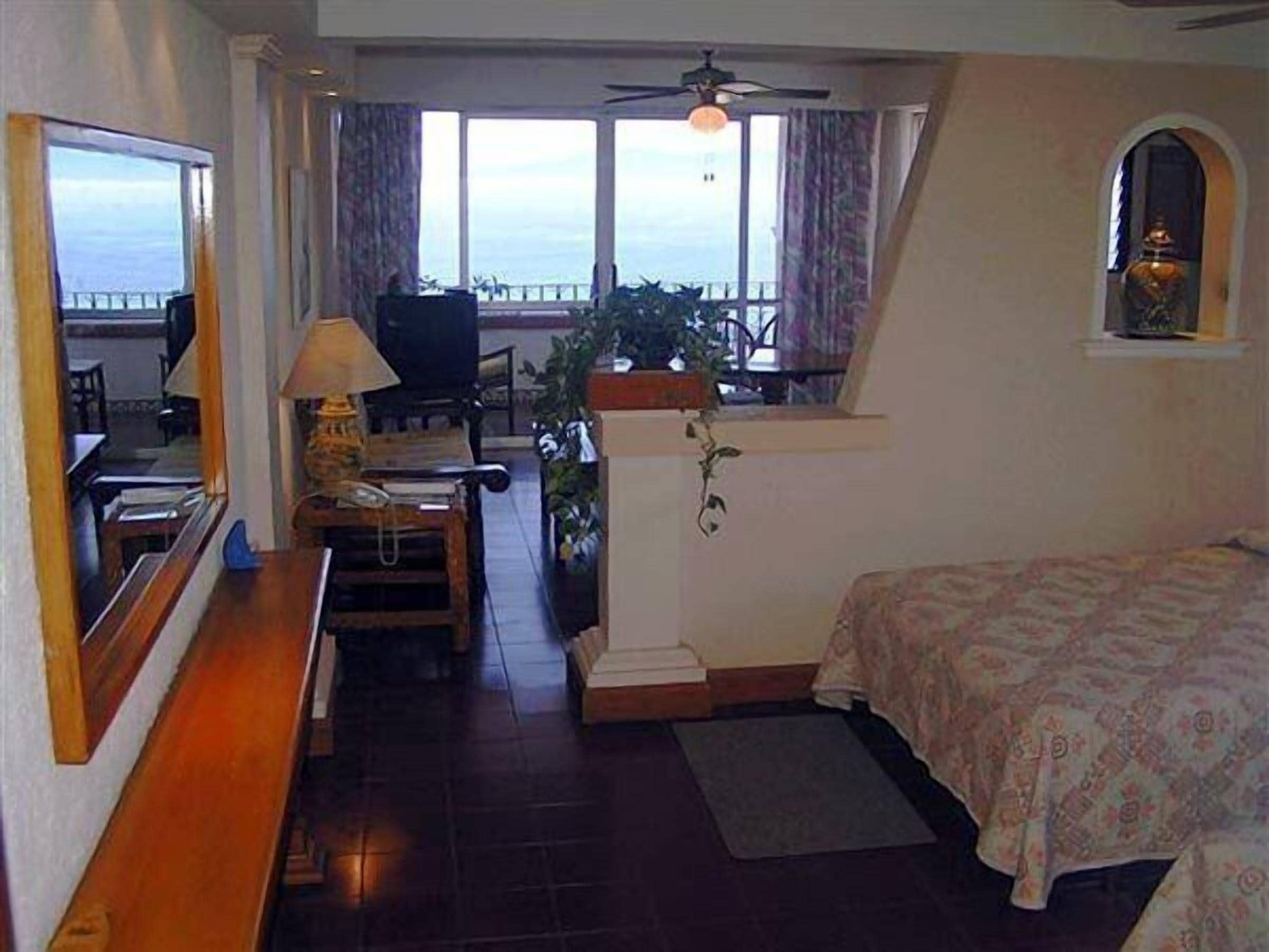 Quarto com cama, escrivaninha, cadeira, abajur, ventoinha no teto e janela com vista para o mar.