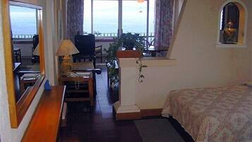 Quarto com cama, escrivaninha, cadeira, abajur, ventoinha no teto e janela com vista para o mar.