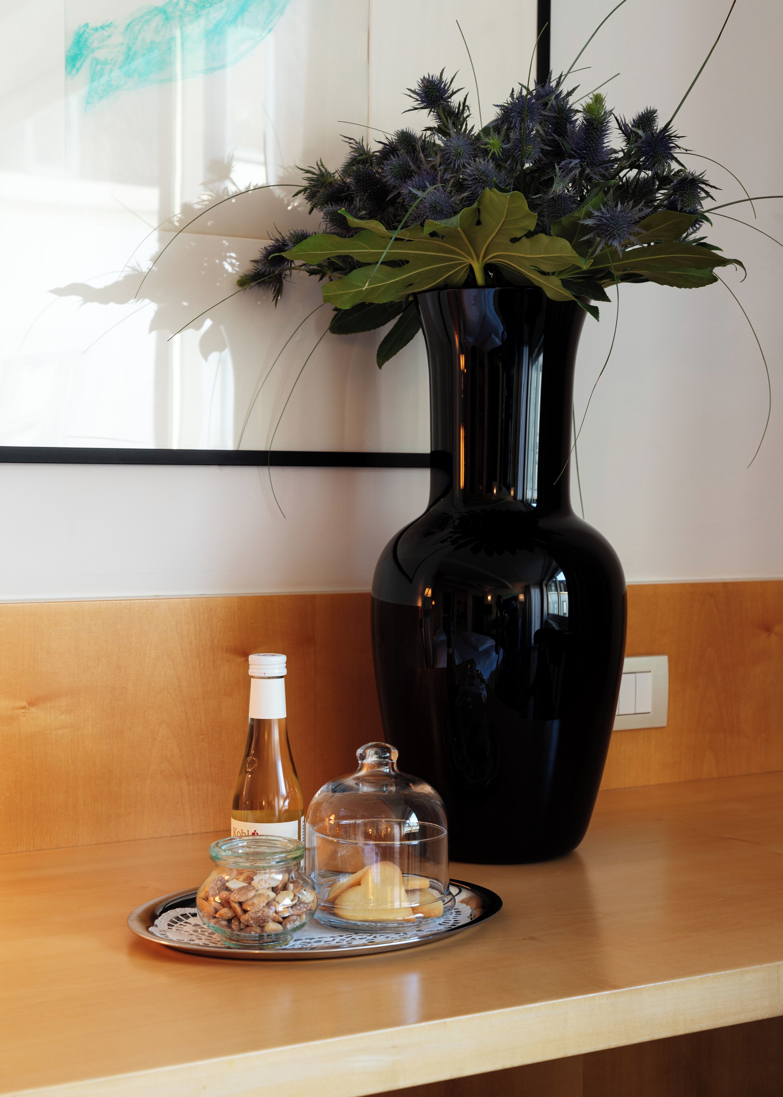 Uma mesa de madeira com um vaso preto contendo um arranjo floral, um pote de vidro com biscoitos e um recipiente de vidro com nozes.