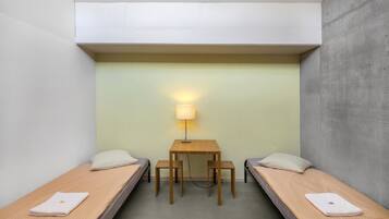 Quarto pequeno e compacto com duas camas de solteiro, uma mesinha e uma luminária.