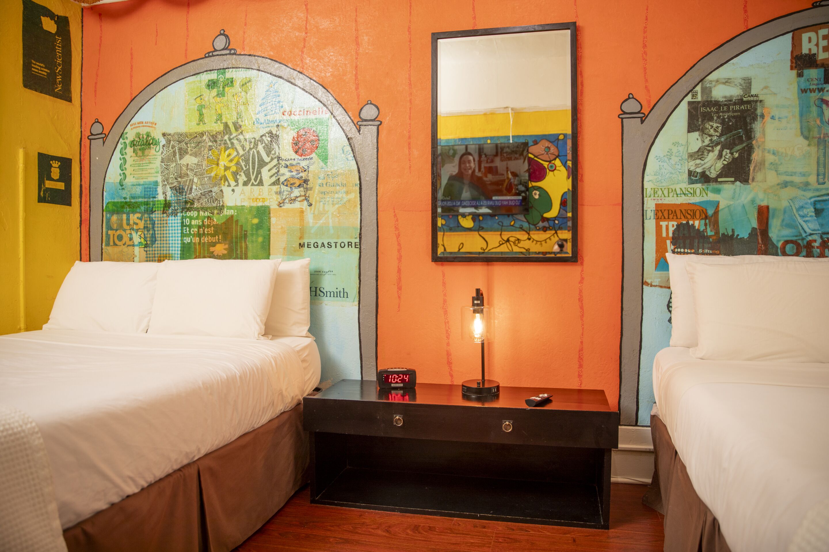 Quarto de hotel com duas camas, uma mesa de cabeceira com abajur, e parede decorada com diversos cartazes e obras de arte.