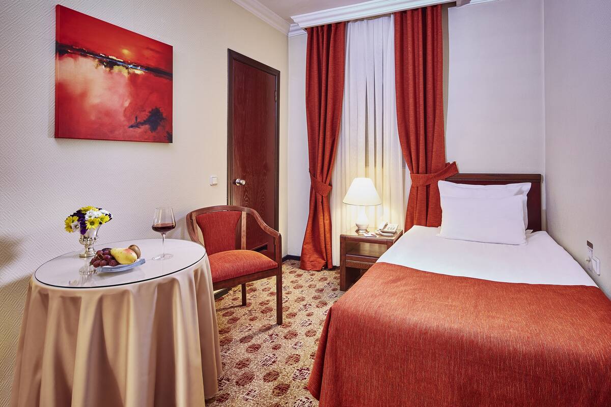Quarto de hotel com uma cama, uma cadeira, uma mesa pequena com um vaso de flores e uma taça de vinho, e um quadro na parede.