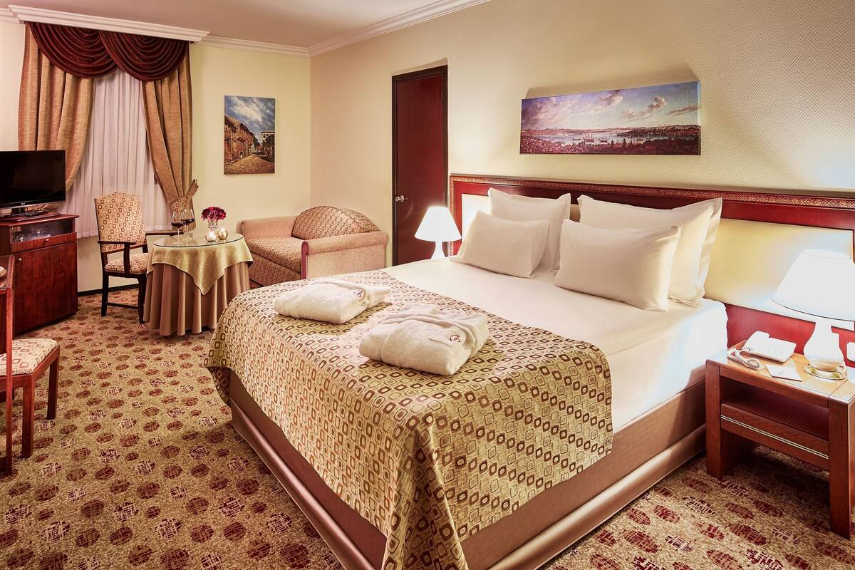 Quarto de hotel com uma cama grande, uma escrivaninha com televisão, uma cadeira e uma mesinha com um vaso de flores.