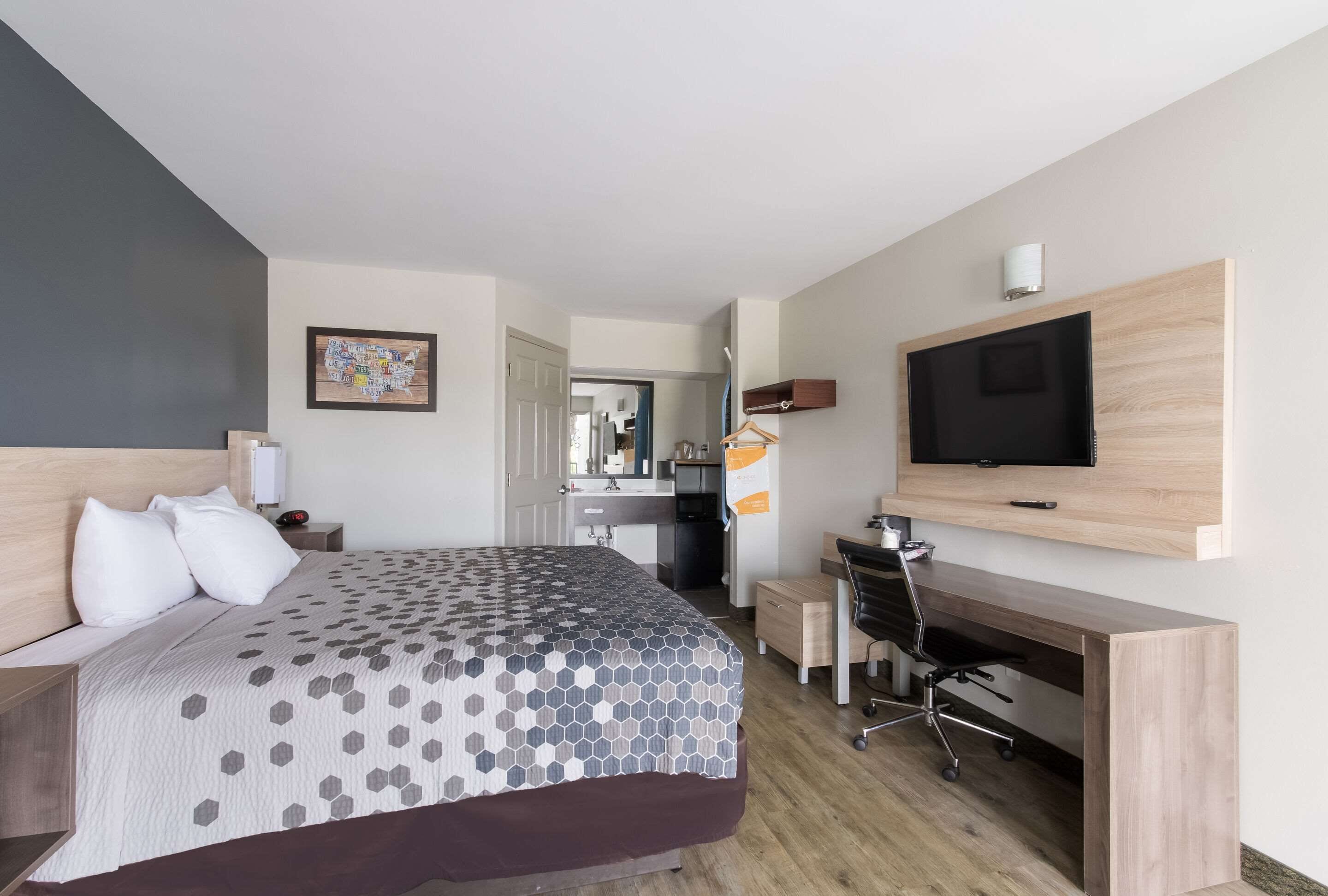 Quarto de hotel com cama, escrivaninha, cadeira, televisão e cozinha compacta.