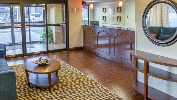 Um saguão de hotel com uma mesa de recepção, área de estar com uma mesa redonda e uma grande janela com vista para a rua.