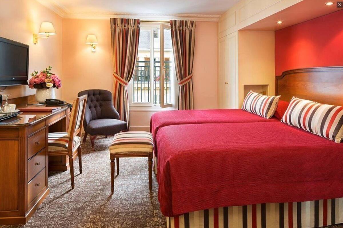 Quarto de hotel com cama, escrivaninha, cadeira, televisão e janela com cortinas.