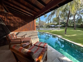 Uma área à beira da piscina com mobiliário de madeira e vista para a piscina e vegetação ao redor.