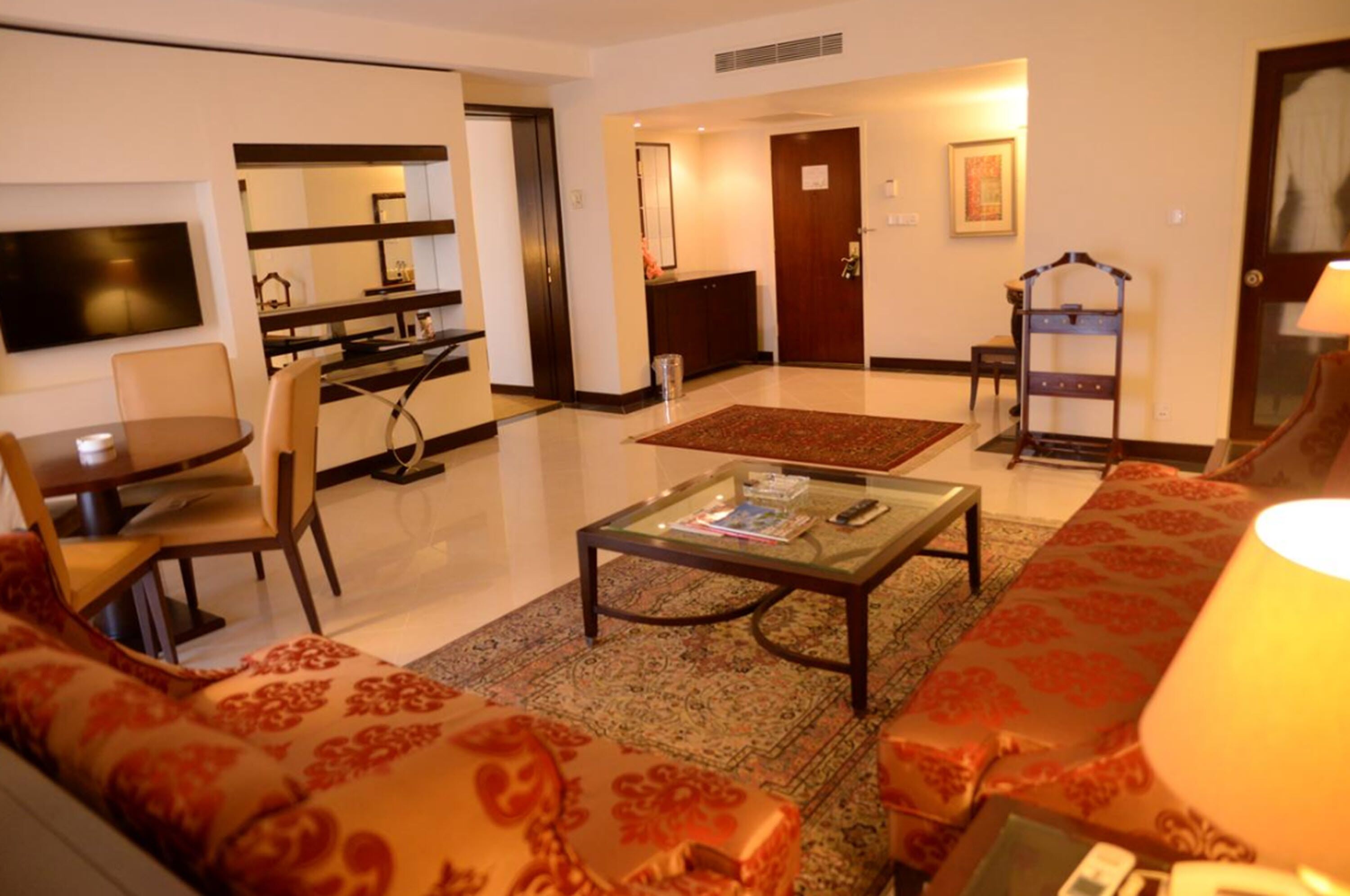 Uma sala de estar moderna com um sofá, mesa de centro e uma TV. Há uma área de jantar com cadeiras e uma mesa. Uma lareira é visível ao fundo.