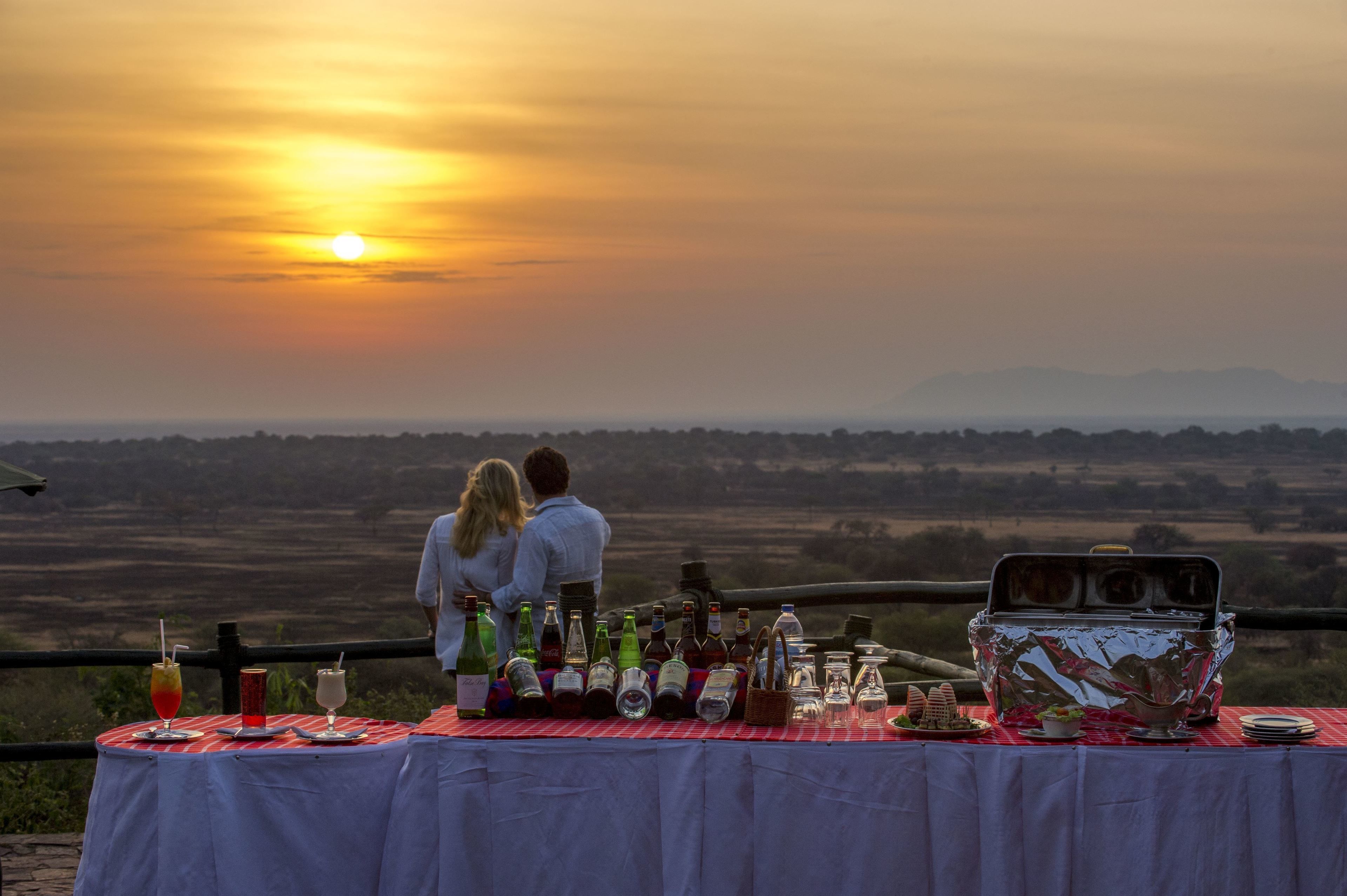 Um casal observa uma vista panorâmica do pôr do sol a partir de uma mesa com várias garrafas e um churrasco.