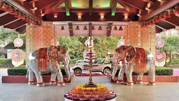Uma entrada decorativa com duas estátuas metálicas de elefante, uma fonte central e um carro branco estacionado ao fundo.