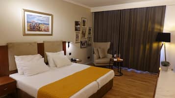 Quarto de hotel com uma cama grande, uma cadeira, uma mesa pequena com abajur e um quadro na parede.