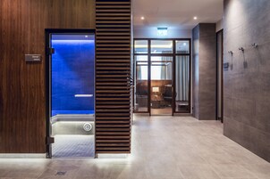 Um hall de hotel moderno com balcão de recepção de madeira, área de sauna iluminada em azul e espaço de estar com luminária e cadeiras.