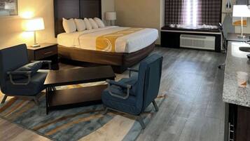 Quarto de hotel com cama, escrivaninha, cadeira, mesa pequena, luminária, radiador e janela com cortinas.
