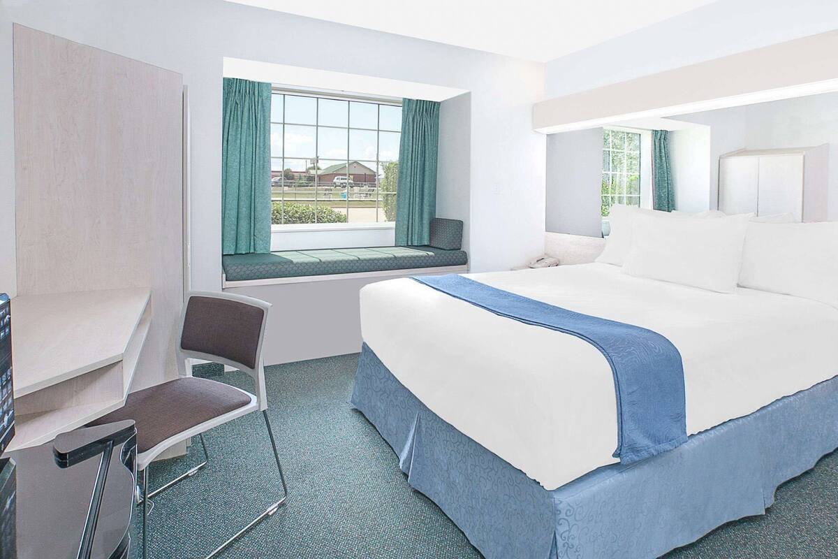 Quarto de hotel com cama, escrivaninha, cadeira e janela com cortinas.