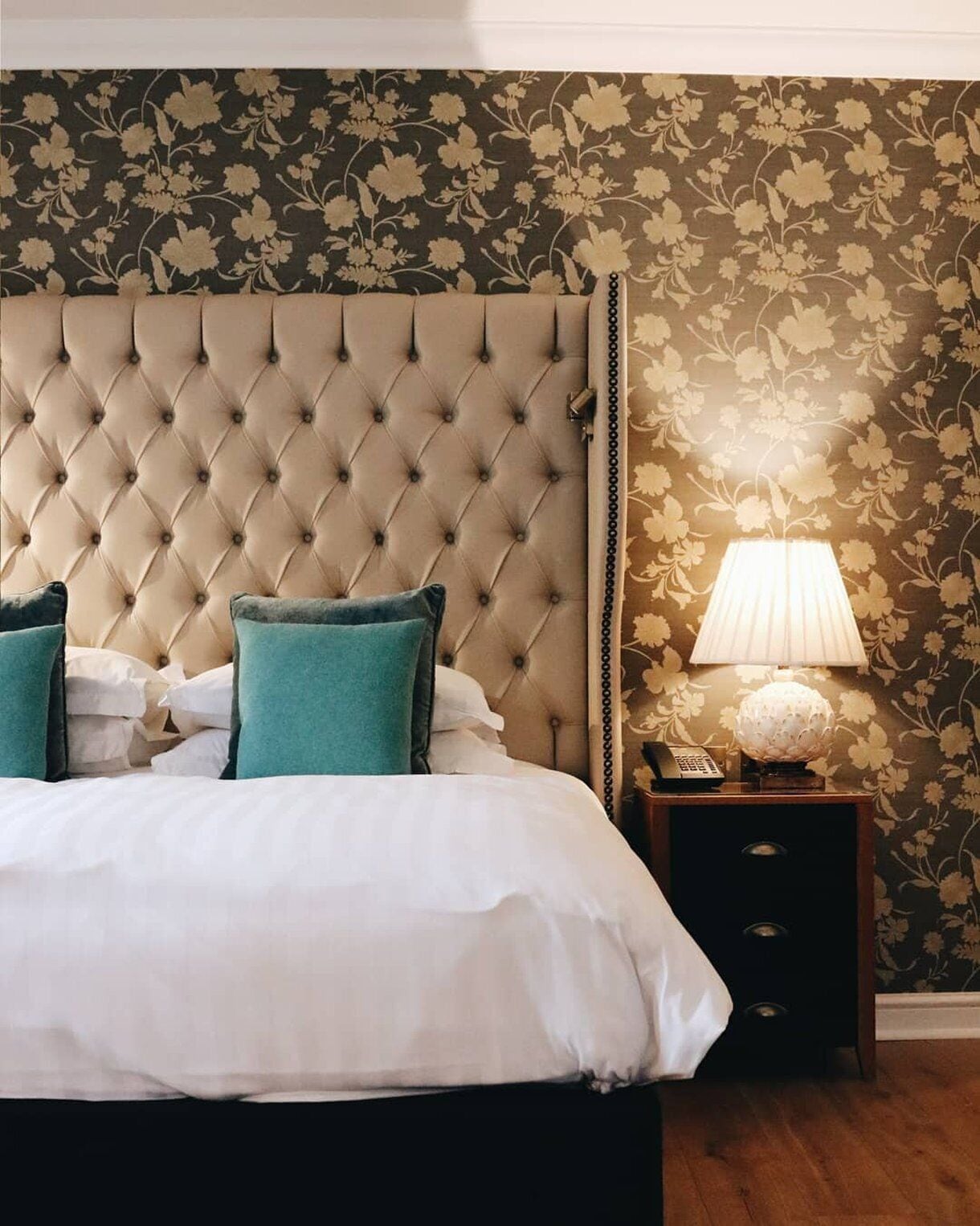 Quarto com cabeceira estofada, cama com roupa de cama branca e almofadas verde-água, mesa de cabeceira com abajur e papel de parede floral.