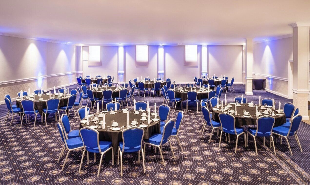 Uma sala de conferências ampla, com mesas redondas e cadeiras azuis dispostas em formato de U.