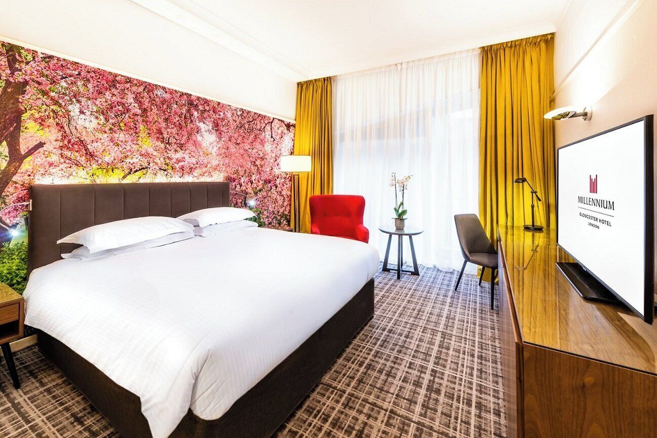 Quarto de hotel com uma cama grande, uma poltrona vermelha, uma mesinha redonda com uma luminária e uma televisão sobre um armário de madeira.