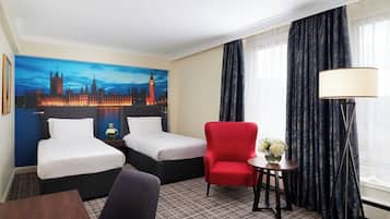 Quarto de hotel com duas camas, uma poltrona vermelha, uma mesa pequena com um vaso e um quadro de uma paisagem urbana na parede.