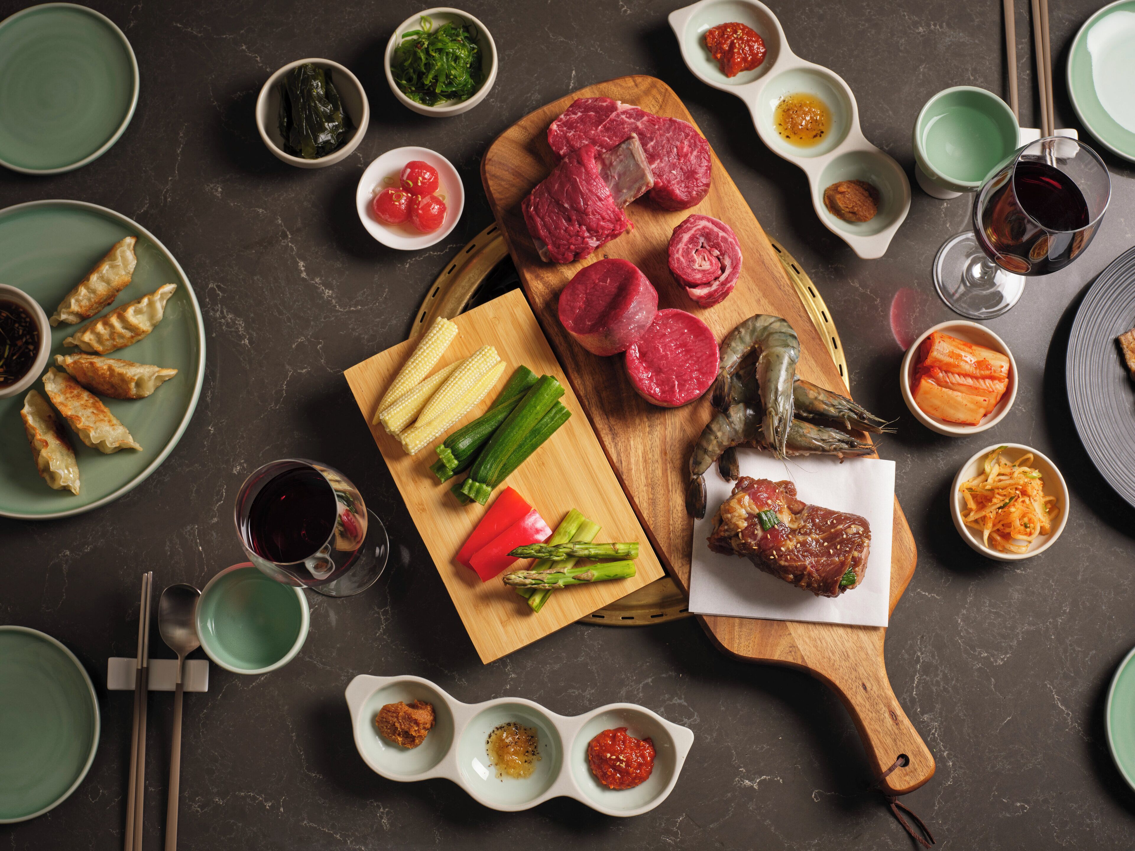 Diversos alimentos como dumplings, milho, aspargos e carne, sobre uma tábua de madeira, acompanhados por pequenos recipientes com molhos e acompanhamentos.