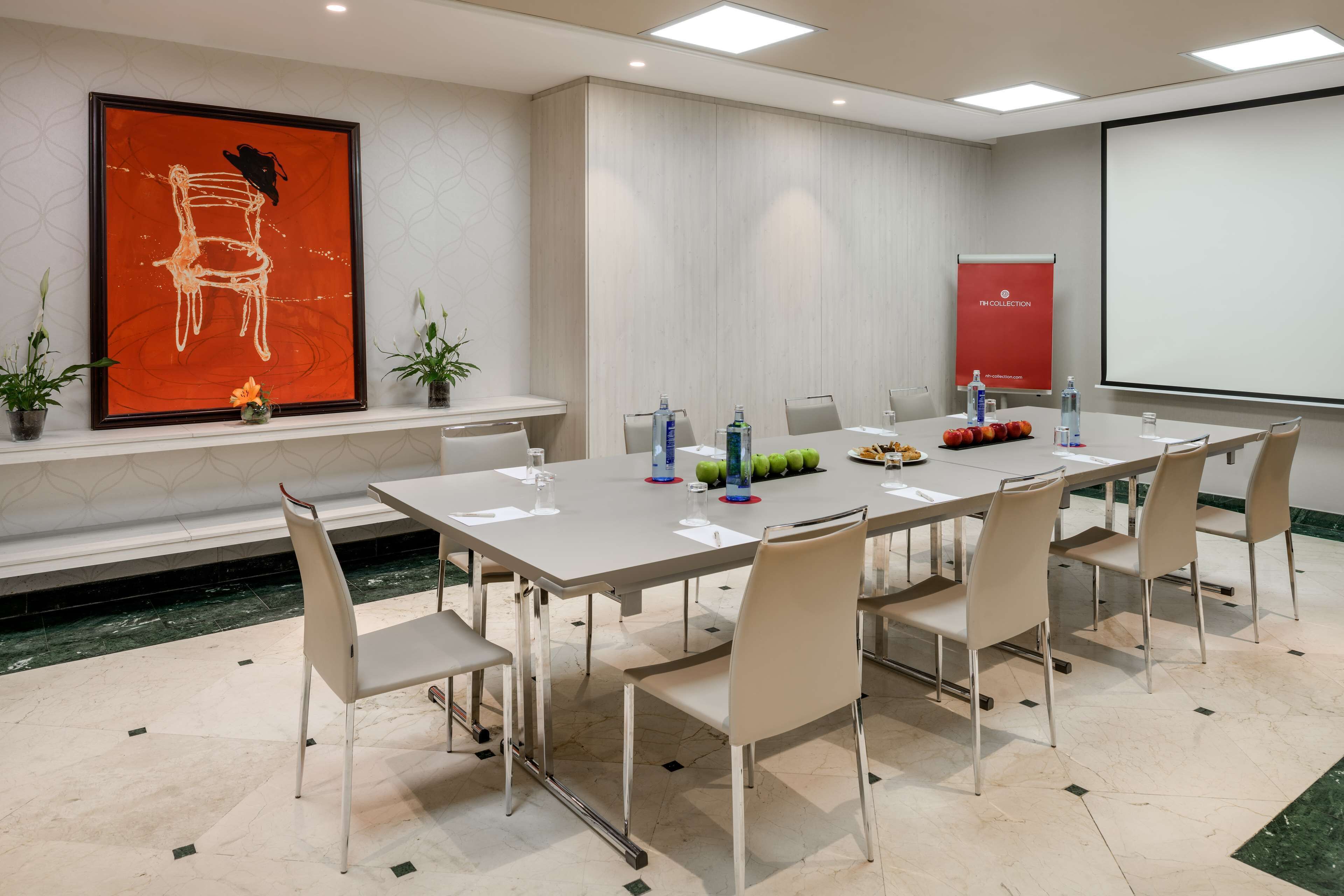 Sala de conferências com uma mesa comprida, seis cadeiras, uma tela de projetor, um banner vermelho e plantas em vasos.