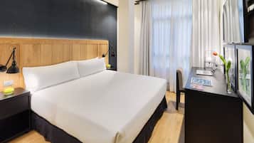 Quarto de hotel com uma cama grande, uma escrivaninha com um laptop, uma mesa de cabeceira com uma luminária e uma janela com cortinas.