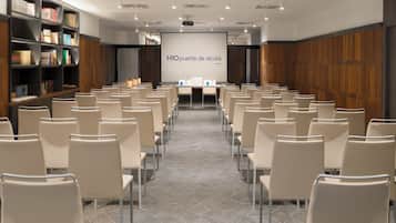 Uma sala de conferências com fileiras de cadeiras, uma tela de projetor e uma estante repleta de livros.