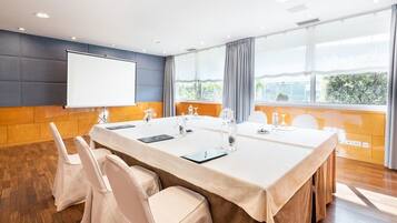 Sala de conferências com uma mesa comprida, cadeiras, tela de projetor e janelas amplas com cortinas.