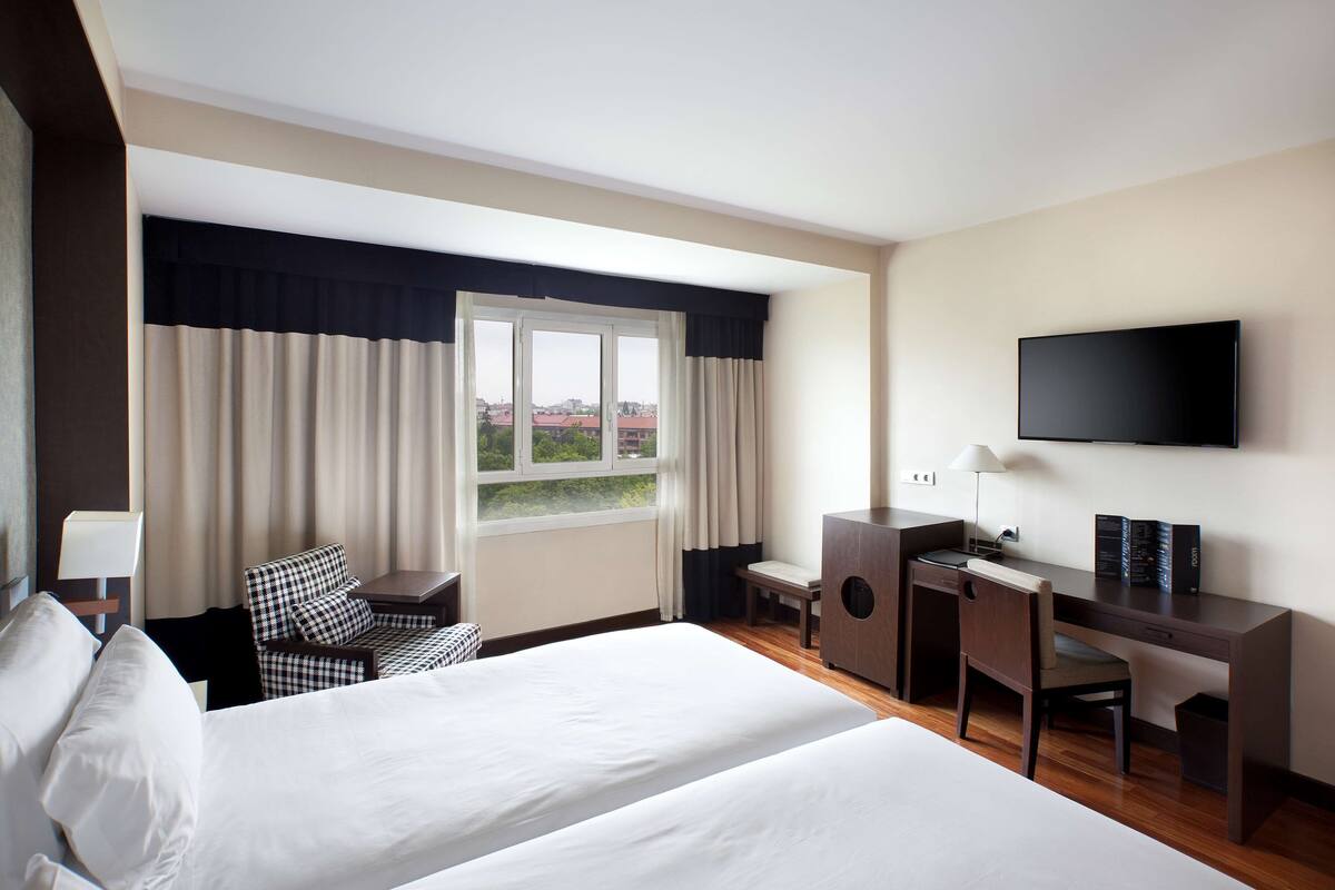 Quarto de hotel com uma cama grande, uma escrivaninha, uma cadeira, uma televisão e uma janela com cortinas.