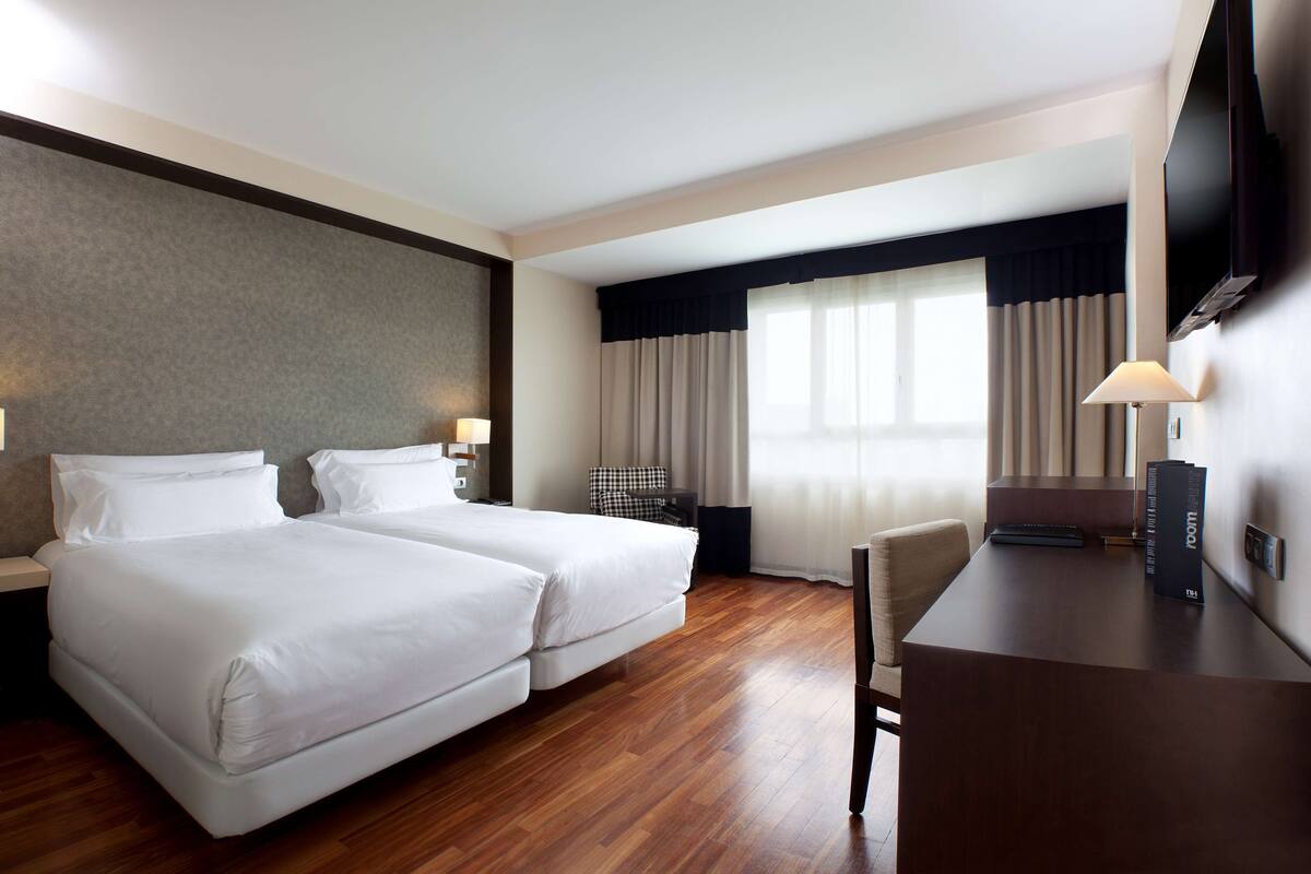 Quarto de hotel com duas camas, uma escrivaninha, uma cadeira, uma televisão e uma janela grande com cortinas.