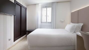 Quarto de hotel moderno com uma cama grande, um guarda-roupa escuro, uma mesa pequena e uma janela com cortinas finas.