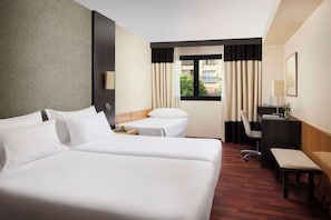 Quarto de hotel com duas camas, uma escrivaninha, uma cadeira e uma janela com cortinas.