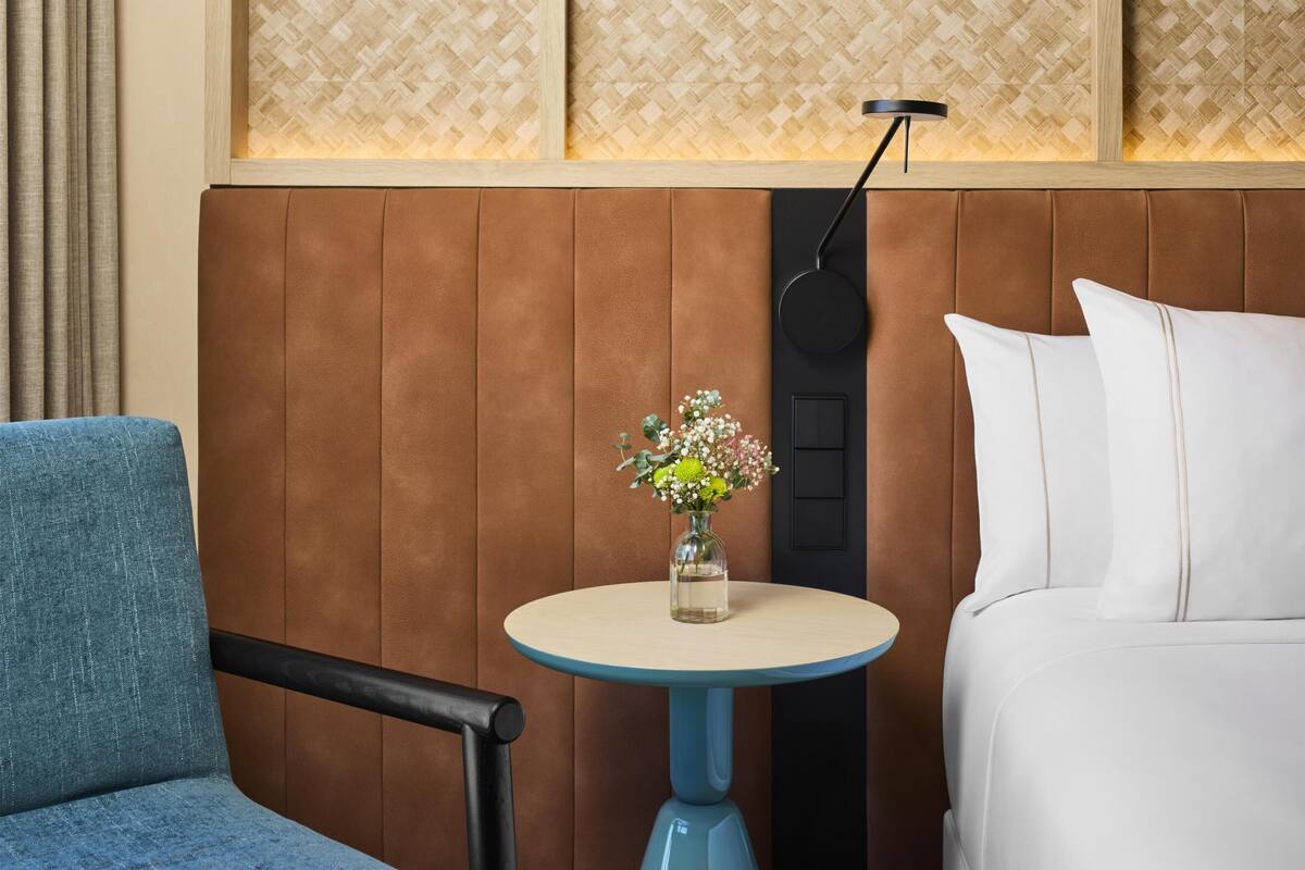 Quarto de hotel com cabeceira marrom, mesa redonda com um vaso de flores, uma cadeira azul e uma cama com almofadas brancas.