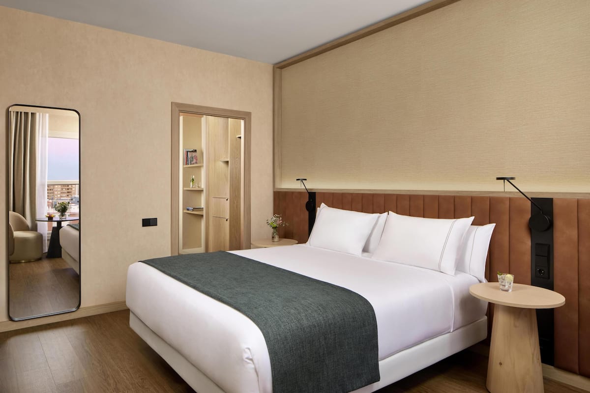 Quarto de hotel com uma cama grande, uma mesa de cabeceira com abajur, um espelho e vista para a cidade pela janela.