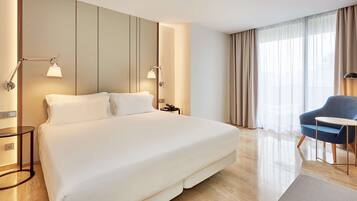 Quarto de hotel moderno com uma cama grande, abajures, uma mesinha e uma poltrona azul.
