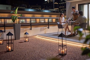 Uma área de refeições no terraço com vista para o horizonte da cidade, incluindo um edifício HSBC. Há mesas e cadeiras, e um garçom está presente.