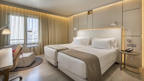 Quarto de hotel com duas camas, uma escrivaninha, uma cadeira, uma luminária e vista para a cidade através de grandes janelas.