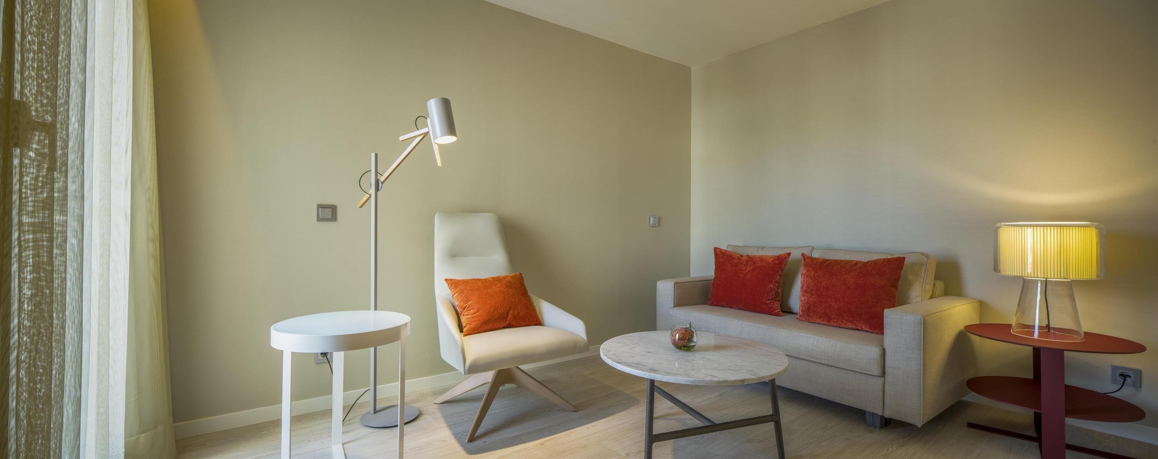 Uma sala de estar moderna com um sofá branco, almofadas laranja, uma mesa de centro redonda, um abajur de chão e um abajur de mesa.