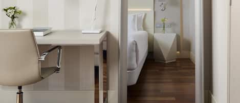 Quarto de hotel moderno com piso de madeira, uma escrivaninha com cadeira, uma cama e uma mesinha de cabeceira com abajur.