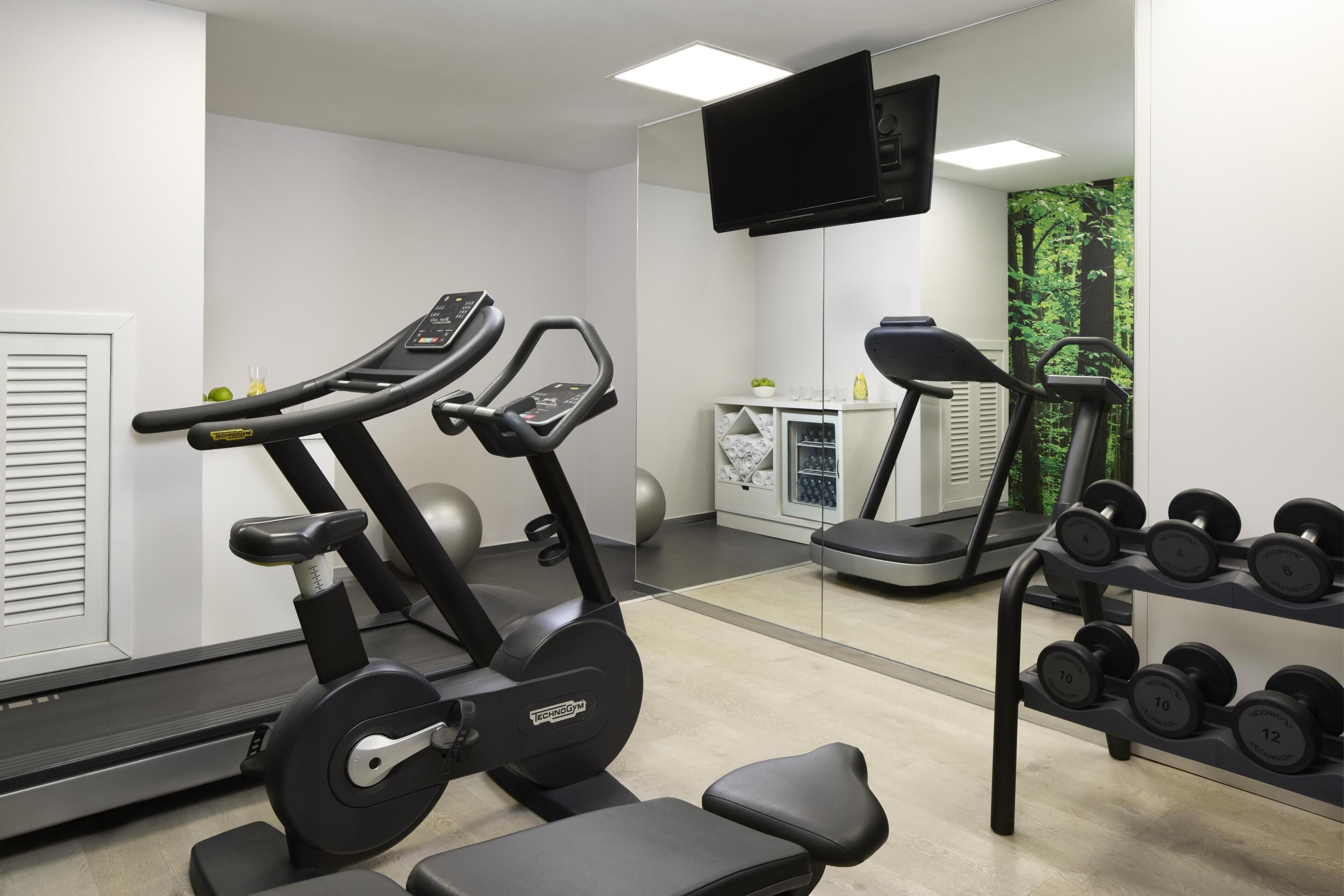 Uma academia com equipamentos de cardio, halteres e uma televisão fixada na parede.