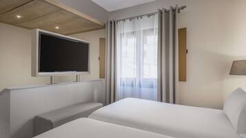 Quarto de hotel com uma cama grande, televisão de tela plana na parede, um banco e uma janela com cortinas.