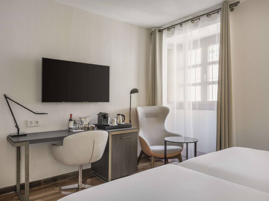 Quarto de hotel com cama, escrivaninha com cadeira, televisão, cafeteira e janela com cortinas.