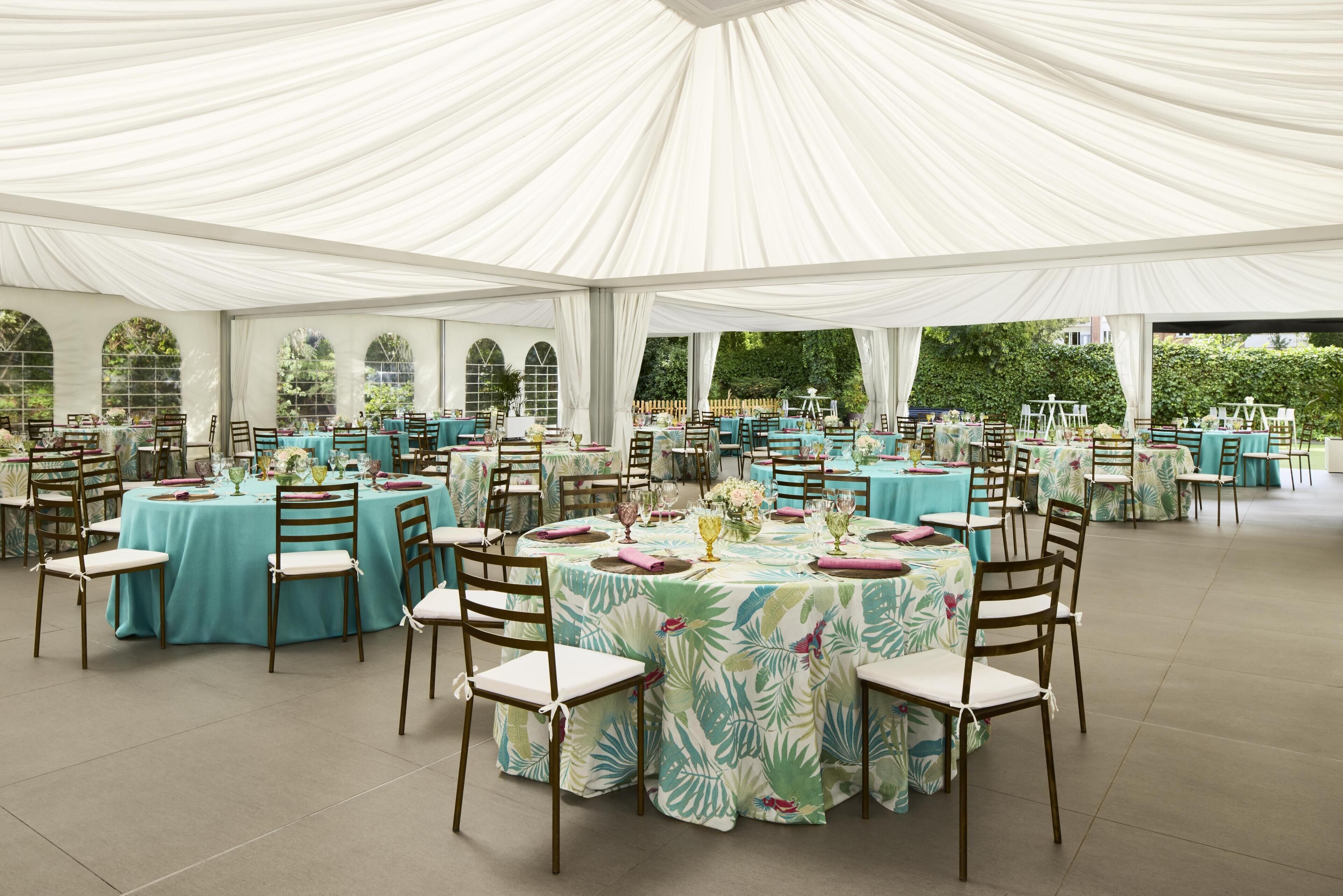 Espaço para eventos ao ar livre com tendas, mesas redondas e cadeiras, coberto por cortinas brancas e toalhas de mesa com padrão de folhagem tropical.