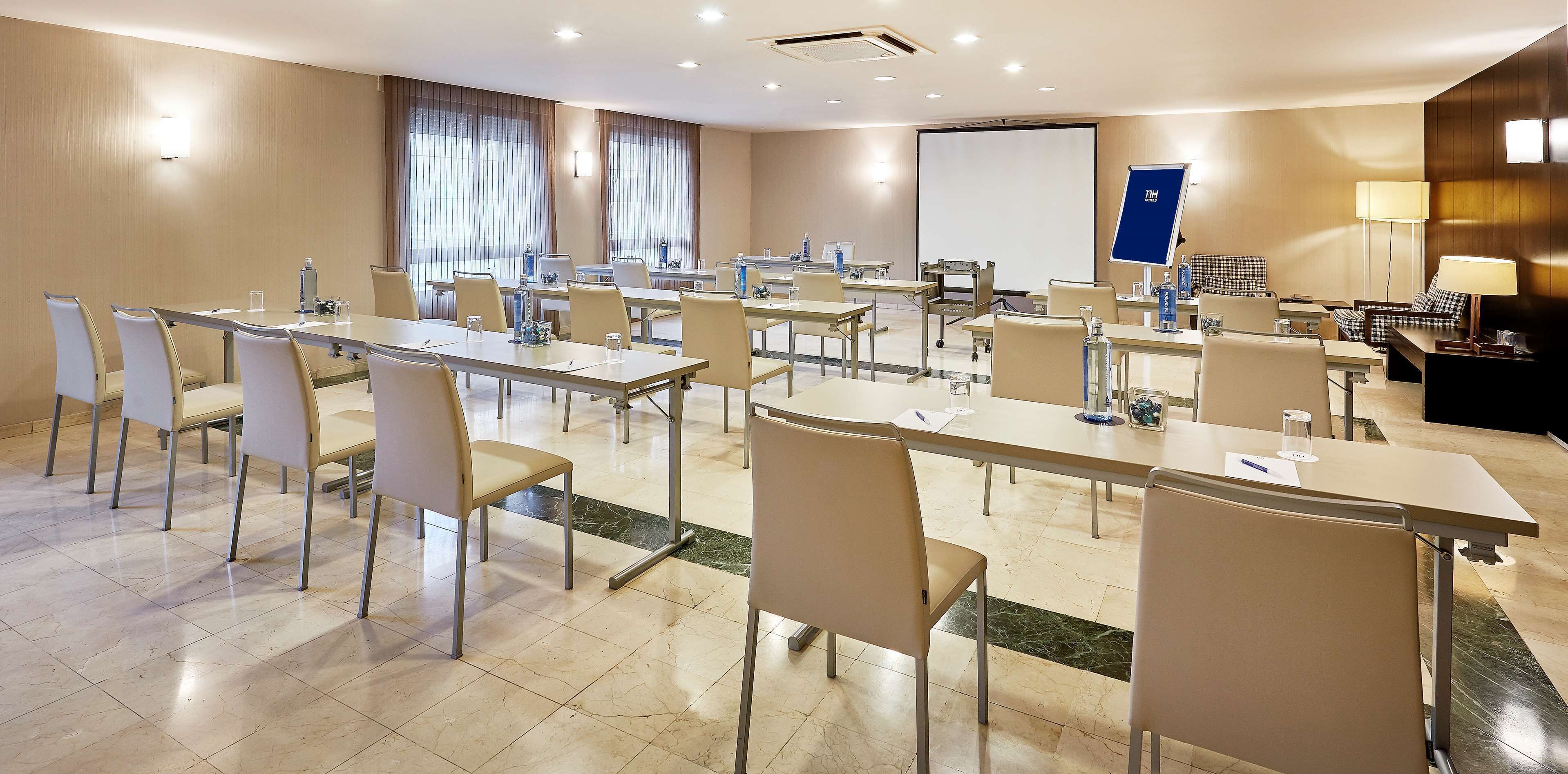 Uma sala de conferências de hotel com várias mesas, cadeiras, uma tela de projetor e um quadro branco.