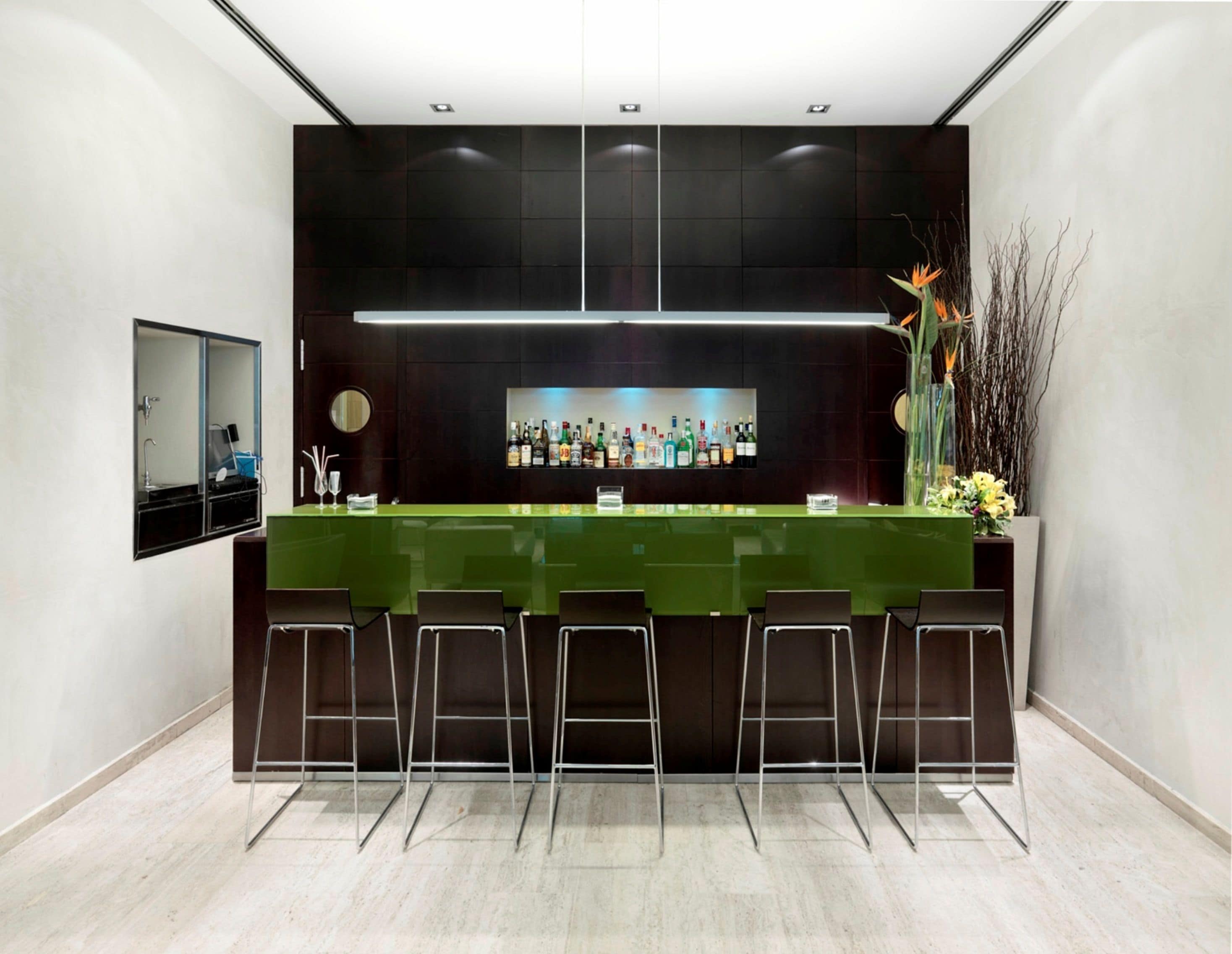 Uma área de bar moderna com bancada verde, vários banquetes e um expositor de bebidas na parede.