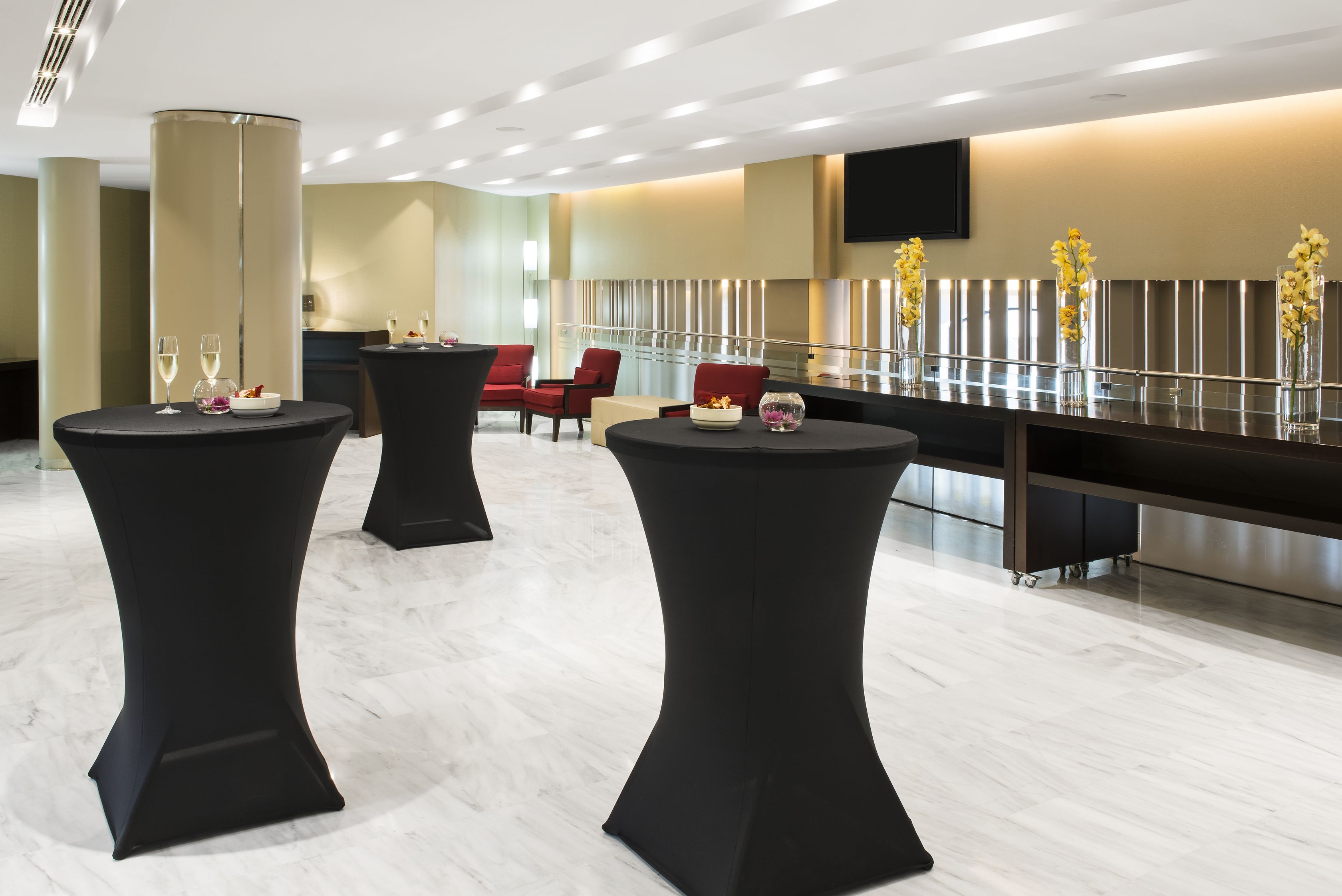 Um lobby de hotel moderno com mesas pretas redondas, cadeiras vermelhas e um balcão de recepção alongado com vasos de flores.