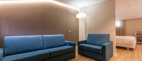 Quarto de hotel moderno com um sofá azul, uma poltrona a combinar, uma mesa de cabeceira e um abajur.